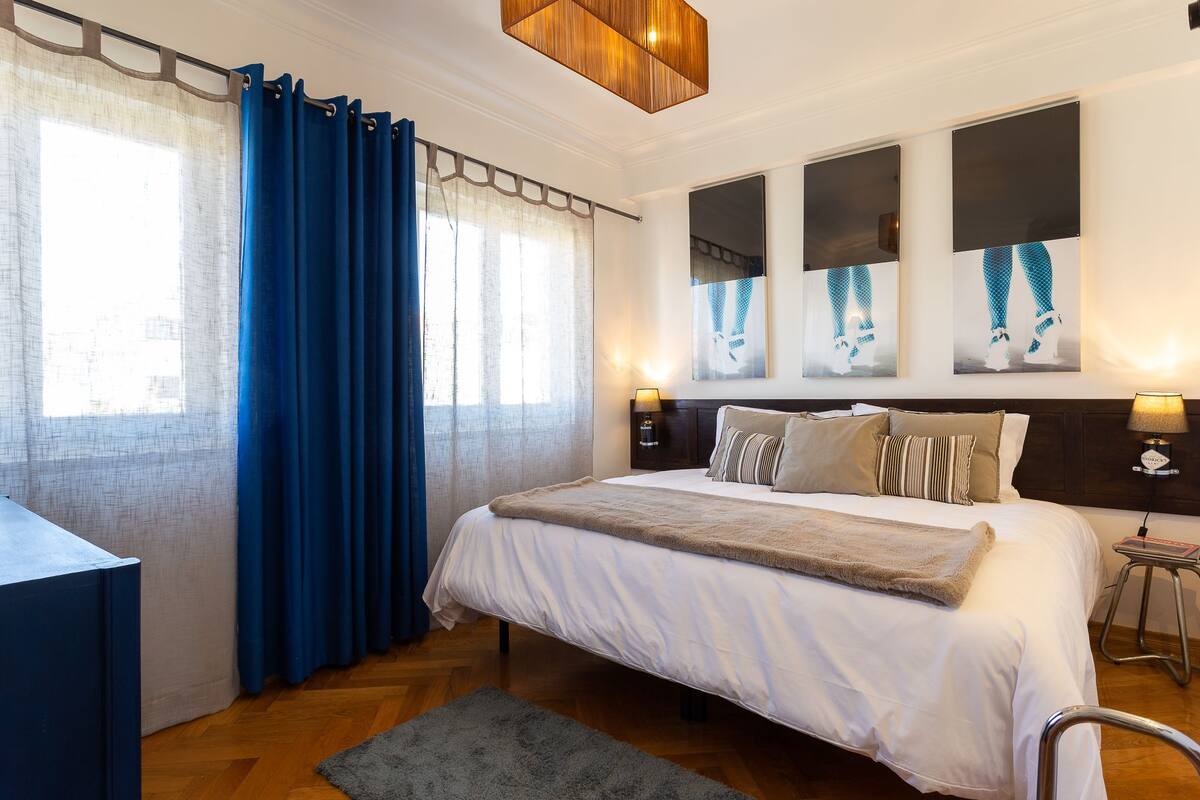 Quarto com cama, mesa de cabeceira, luminária e janela com cortinas.