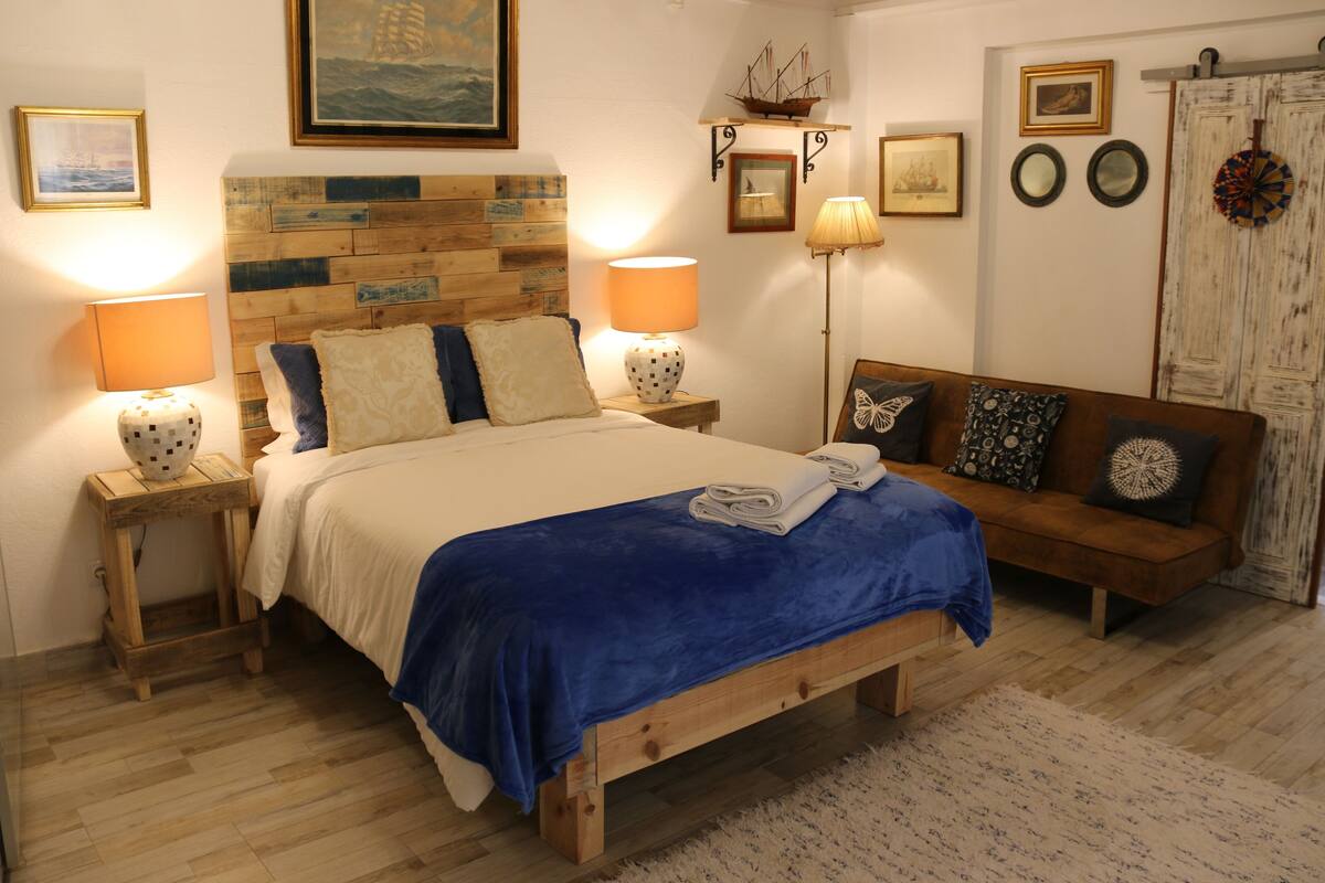 Quarto com cabeceira de madeira, cama com cobertor azul, sofá marrom e duas mesas de cabeceira com luminárias.