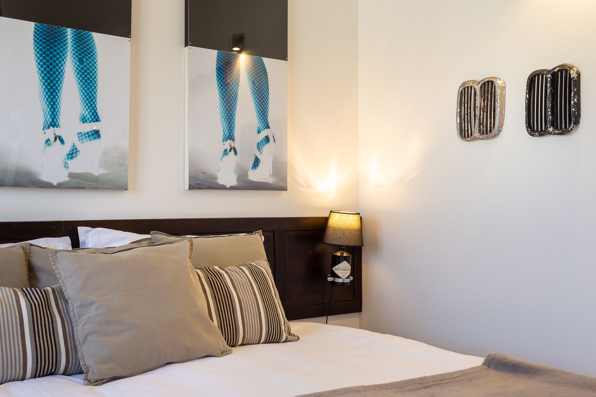 Quarto de hotel com cama, almofadas, cabeceira, luminária de parede e duas obras de arte.