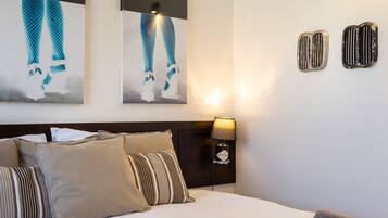 Quarto de hotel com cama, almofadas, cabeceira, luminária de parede e duas obras de arte.