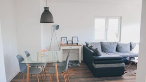 Uma sala de estar moderna com mesa de jantar de vidro, cadeiras cinzentas, sofá escuro com almofadas e uma escrivaninha com computador e quadros.