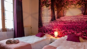 Quarto de hotel com duas camas, cada uma com colcha acolchoada rosa e lençóis brancos. Há uma janela com cortinas, um radiador e um mural de flores e árvores em tons de rosa.
