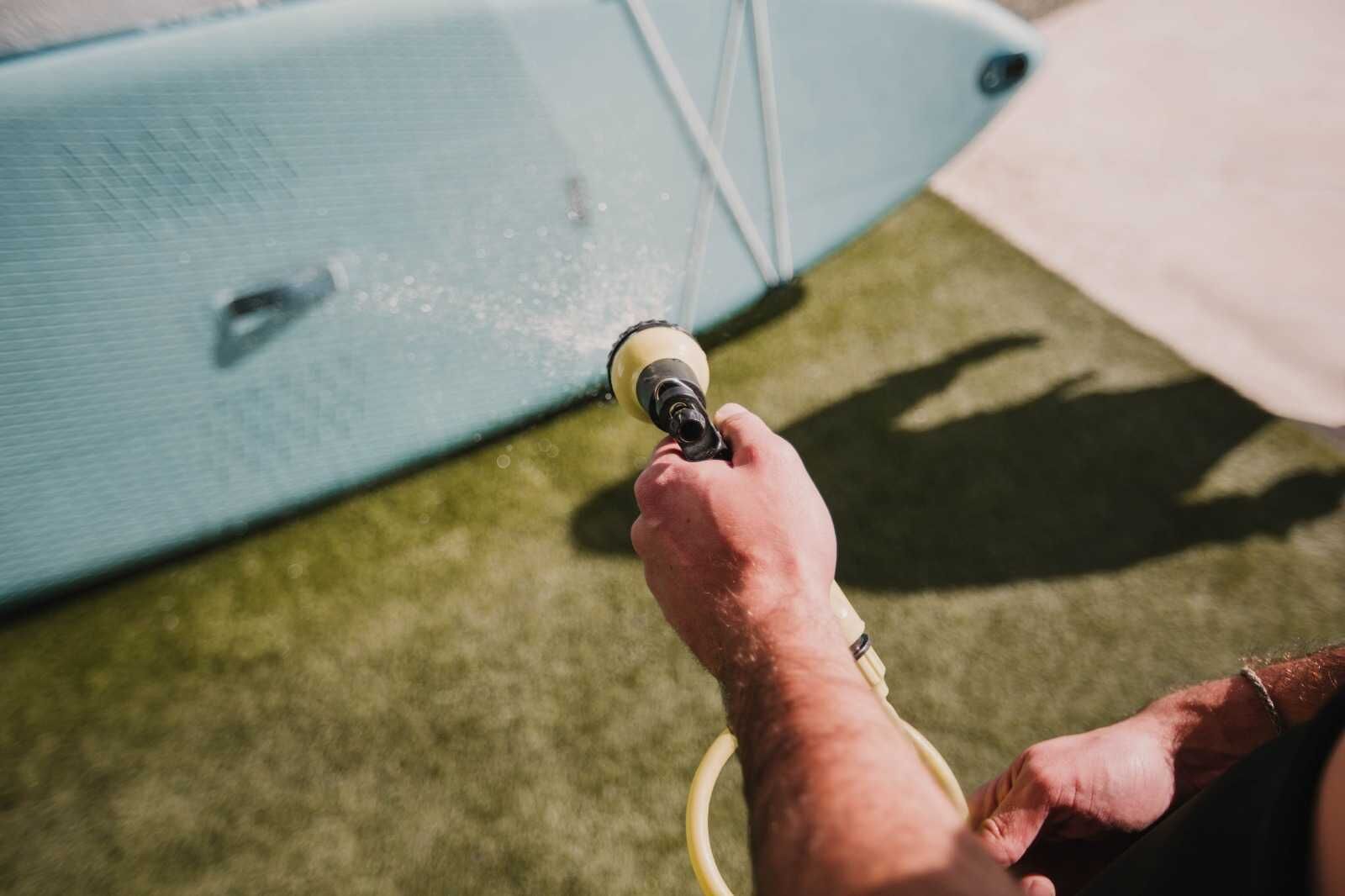 Mão segurando uma mangueira, borrifando água em uma prancha de surf.