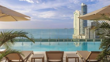 Uma área à beira da piscina com espreguiçadeiras e guarda-sóis, com vista para um mar azul e o horizonte da cidade.