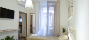 Quarto com cama, televisão fixada na parede, mesa de cabeceira com planta e janela com cortinas leves.