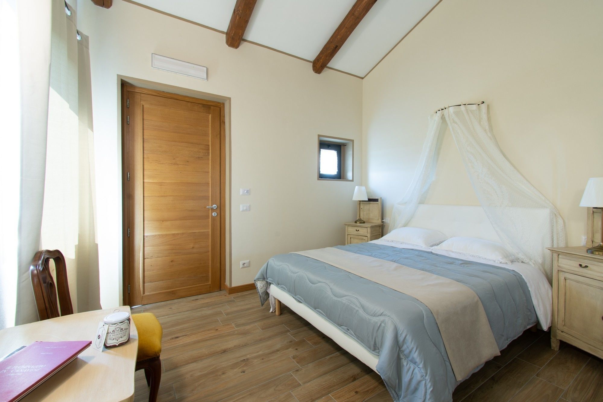 Um quarto com cama, mesas de cabeceira, uma porta de madeira, uma janela pequena e uma cadeira.