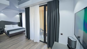 Quarto de hotel moderno com uma cama grande, uma escrivaninha com luminária e uma porta de vidro deslizante para uma varanda.