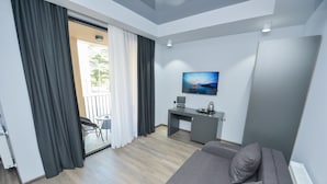 Quarto moderno com sofá cinzento, televisão de tela plana, escrivaninha e varanda com cortinas.