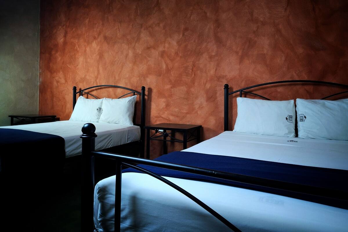 Duas camas de solteiro com estrutura de metal e roupa de cama branca em um quarto com parede laranja texturizada.