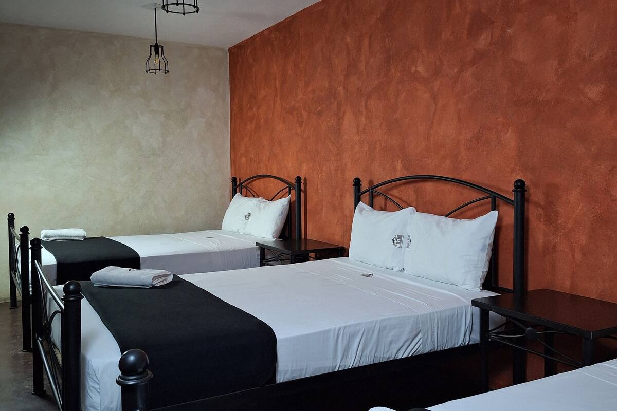 Quarto de hotel com três camas de solteiro, cada uma com estrutura preta e roupa de cama branca. Há uma mesa de cabeceira de madeira com luminária entre as camas. O quarto possui parede texturizada marrom e teto branco liso.