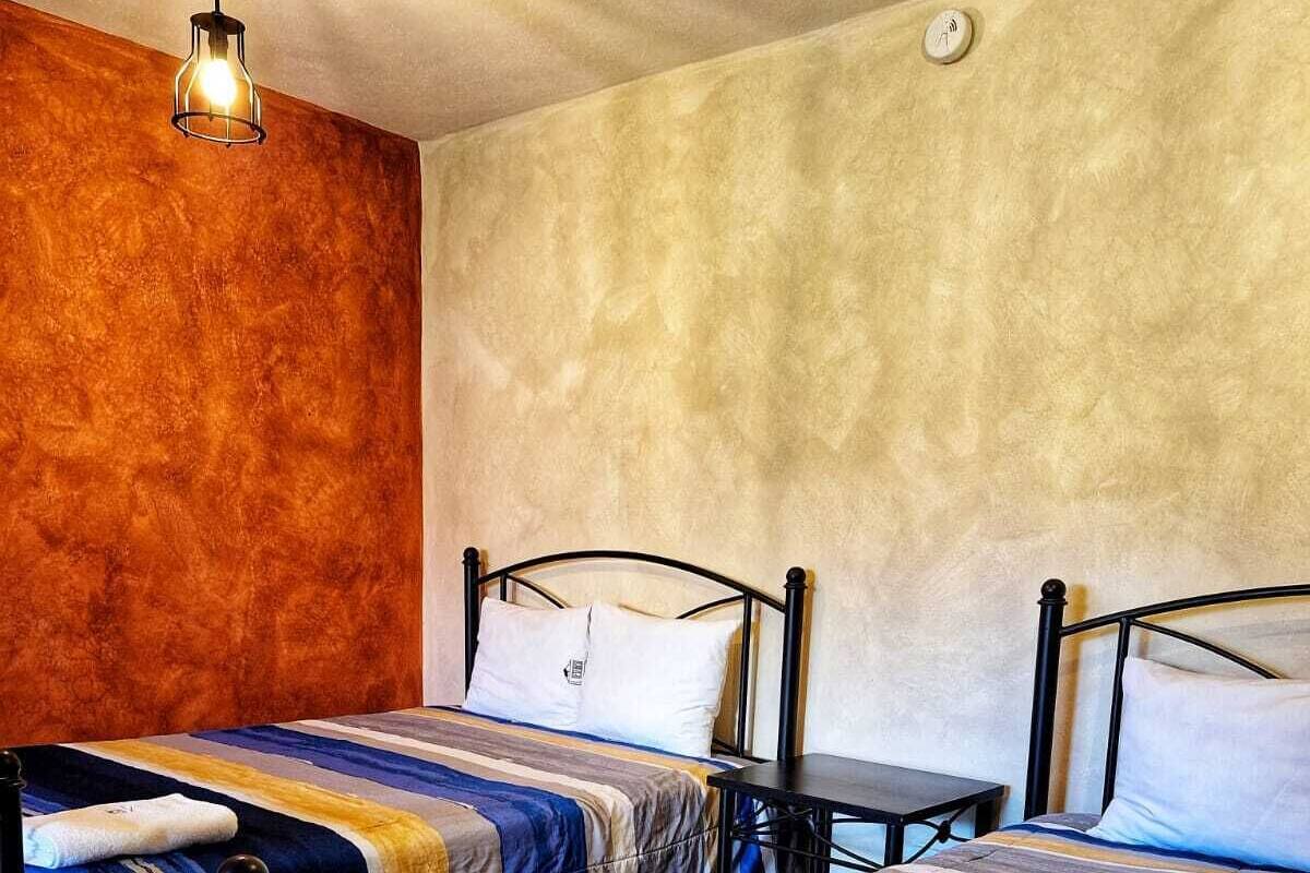 Quarto com duas camas, cada uma com estrutura de metal e roupa de cama listrada. Há uma pequena mesa entre as camas, e uma luminária suspensa sobre ela.