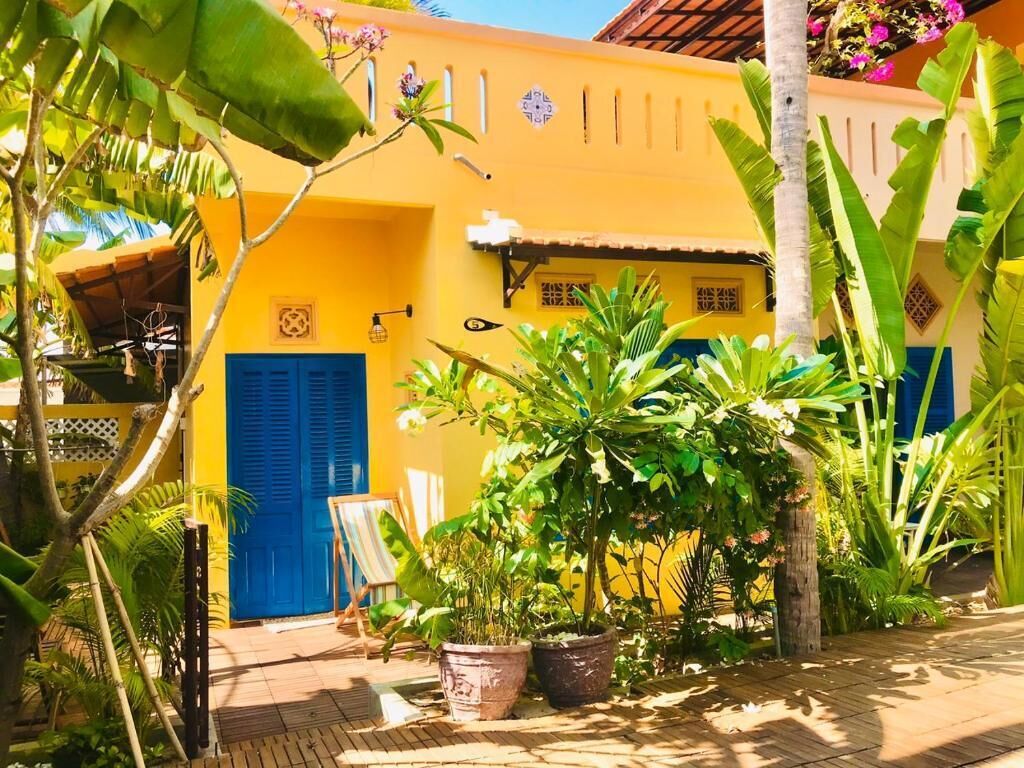 Um edifício amarelo com persianas azuis, rodeado por plantas tropicais e um alpendre de madeira.