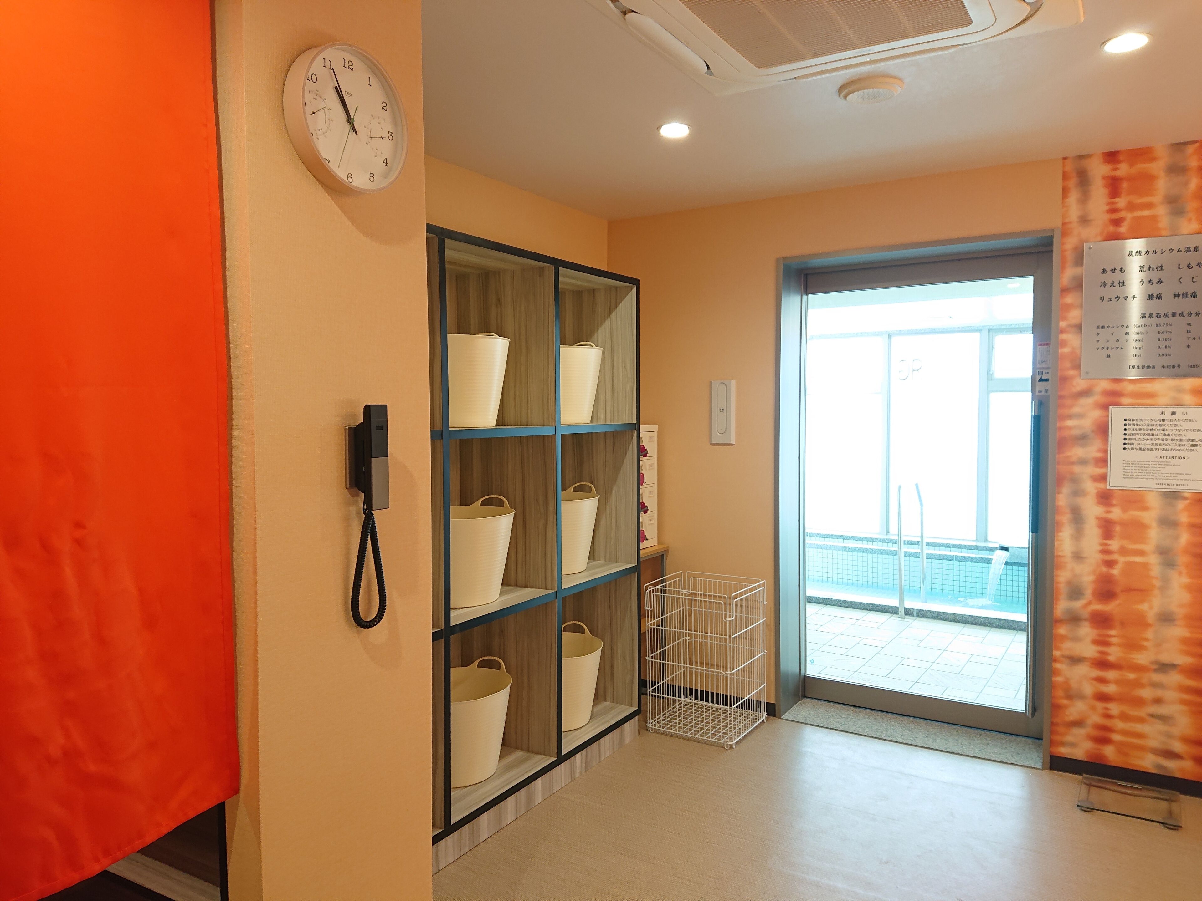 Uma área de recepção de hotel com relógio, telefone, prateleira com cestos de armazenamento e porta que dá para o exterior.