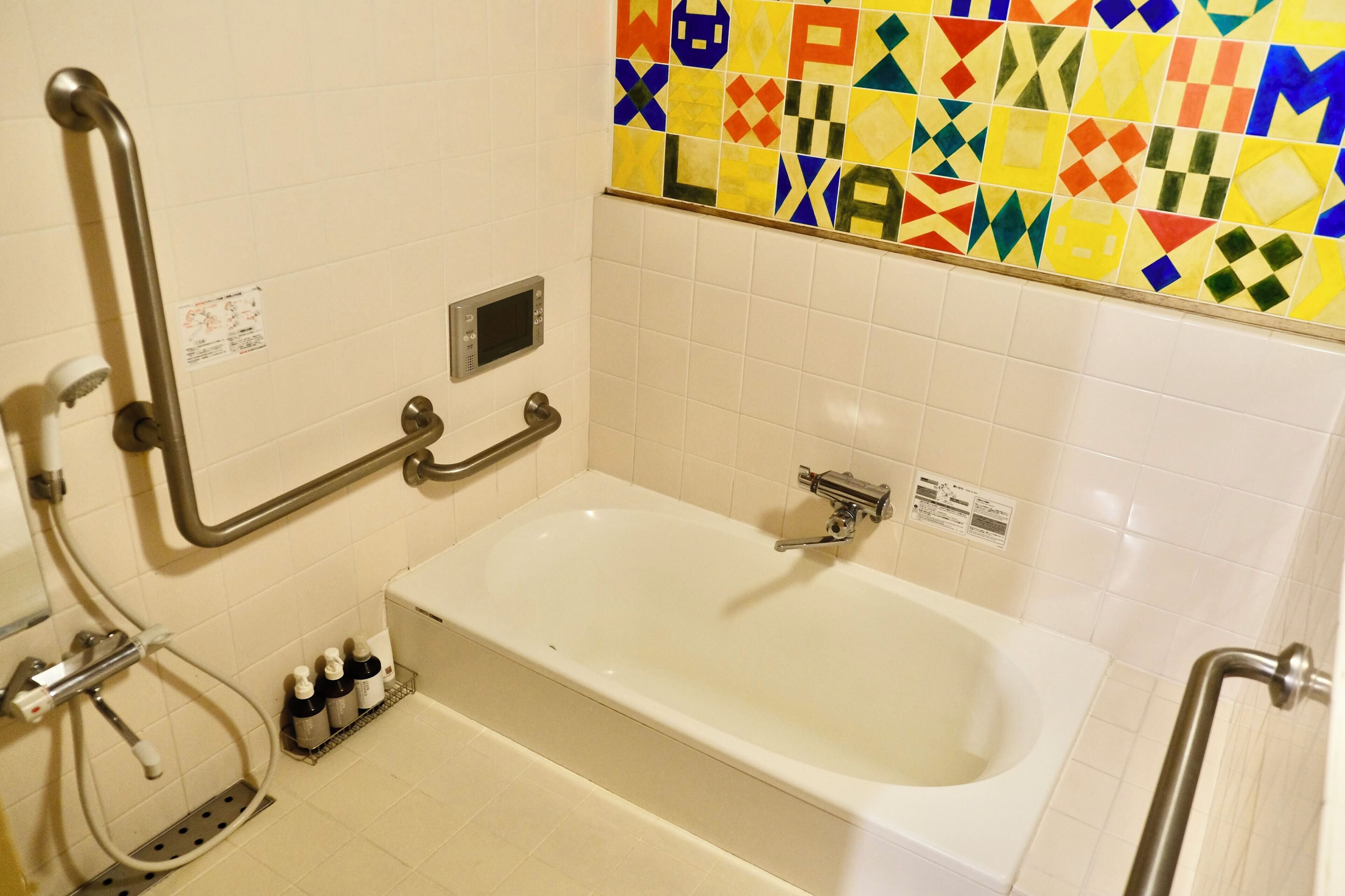 Casa de banho com banheira branca, barras de apoio, chuveiro e parede com mosaico colorido.