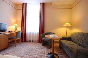 Quarto de hotel com uma escrivaninha, televisão, sofá, mesa redonda pequena e uma janela com cortinas.