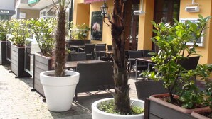 Área externa com assentos, vasos de plantas e cadeiras, um edifício amarelo com uma placa, e calçada de tijolos.