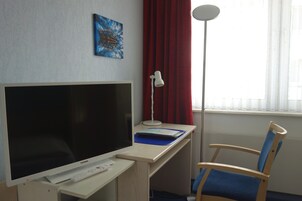 Quarto de hotel com uma televisão de ecrã plano grande, uma secretária com candeeiro, uma cadeira e uma janela com cortinas.