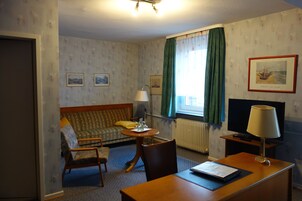 Quarto de hotel com sofá, poltrona, mesa redonda pequena, escrivaninha, luminária, televisão e quadro na parede.