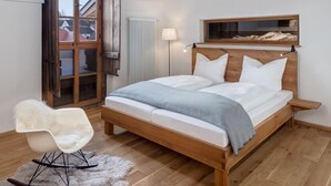 Quarto com cama de madeira, cadeira branca, tapete de pele e janela com vista para edifícios.
