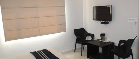 Um quarto com uma cama, uma escrivaninha, duas cadeiras, uma televisão e uma janela com persianas.