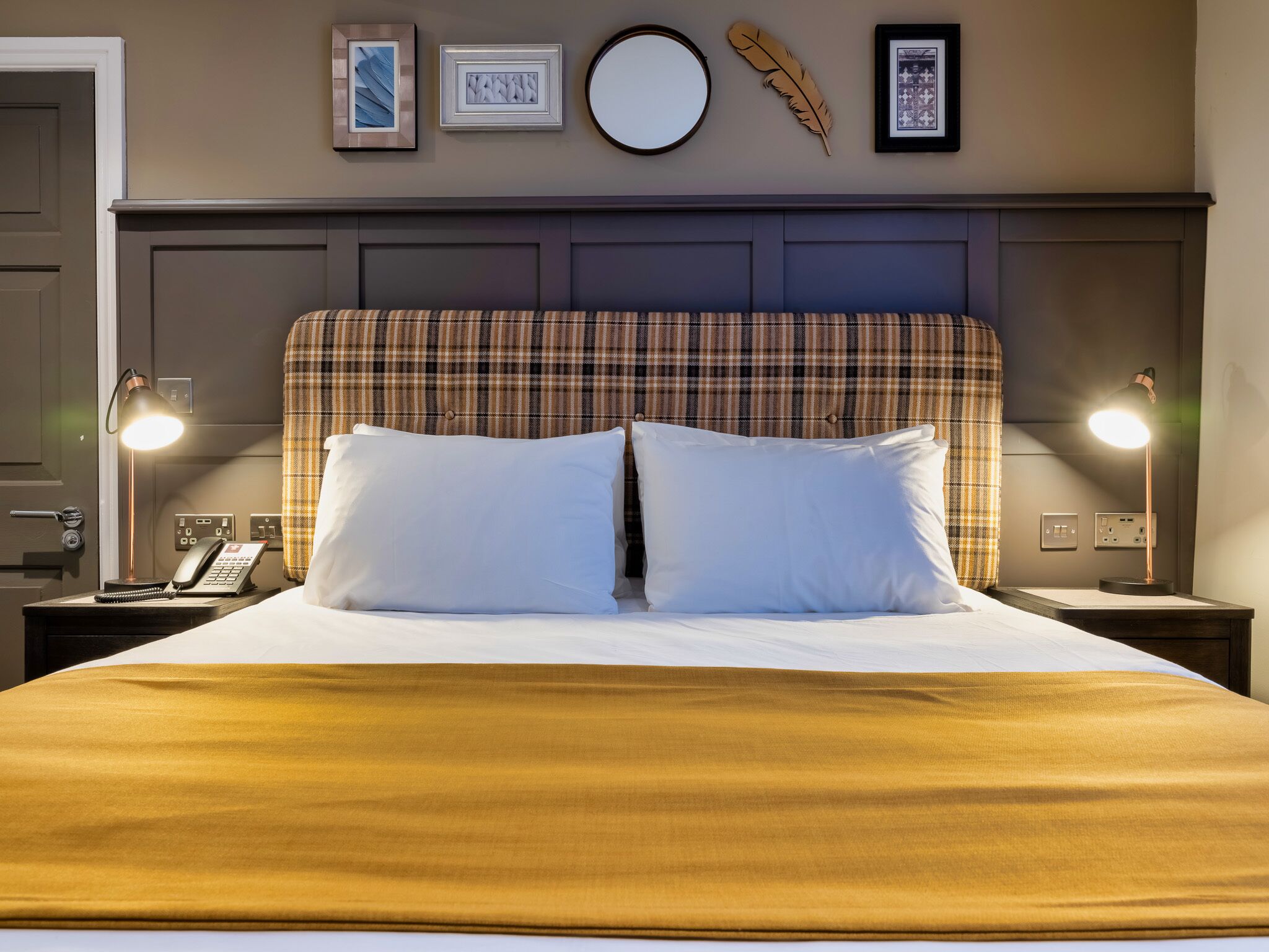 Cama bem arrumada com travesseiros brancos e um cobertor amarelo-mostarda, com duas luminárias de cabeceira, em um quarto com cabeceira xadrez e obras de arte emolduradas na parede.