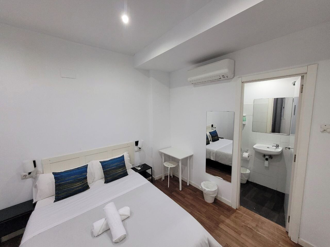 Quarto de hotel com cama, escrivaninha, espelho, pia e um pequeno cesto de lixo.