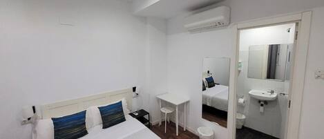 Quarto de hotel com cama, escrivaninha, espelho, pia e um pequeno cesto de lixo.