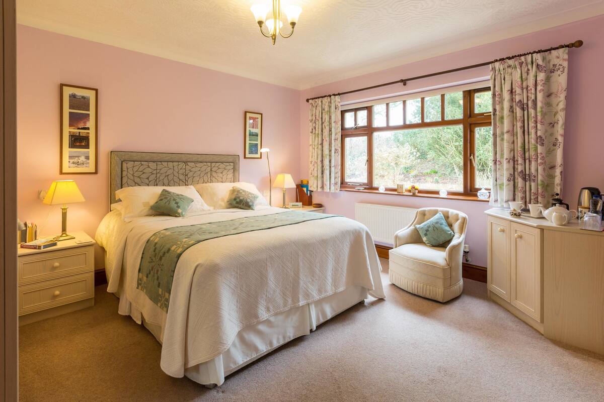 Quarto com cama grande, mesa de cabeceira, cadeira, cômoda e janela com cortinas.
