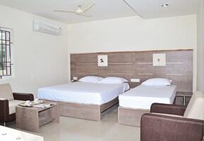 Quarto de hotel com duas camas, um ventilador de teto, uma janela com grades e uma pequena mesa com revistas.