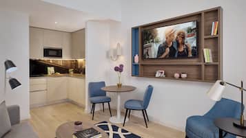 Uma sala de estar moderna com um sofá, uma mesa de centro e duas cadeiras azuis. Há uma televisão presa à parede acima de uma prateleira de madeira com livros e objetos decorativos.