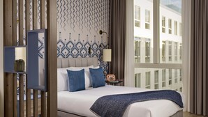Quarto de hotel com cama, mesa de cabeceira, luminária e vista para prédios pela janela.