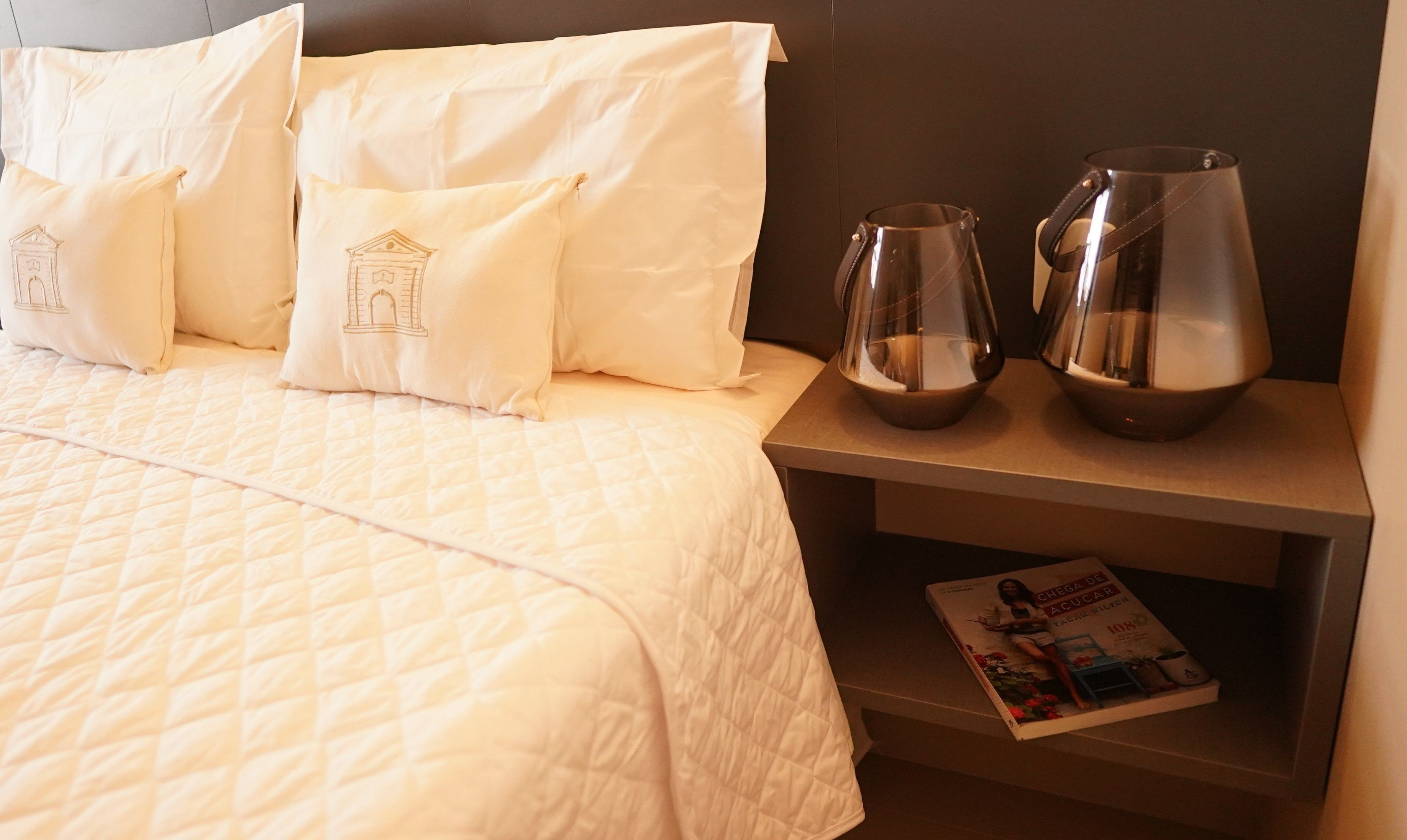 Uma cama bem arrumada com edredom branco acolchoado, uma mesinha de cabeceira com dois jarros de aço inoxidável e uma revista na estante.