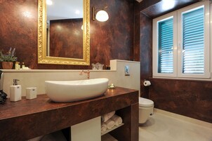 Um banheiro com uma pia oval grande, um espelho alto com moldura dourada, um vaso sanitário e uma janela com persianas.