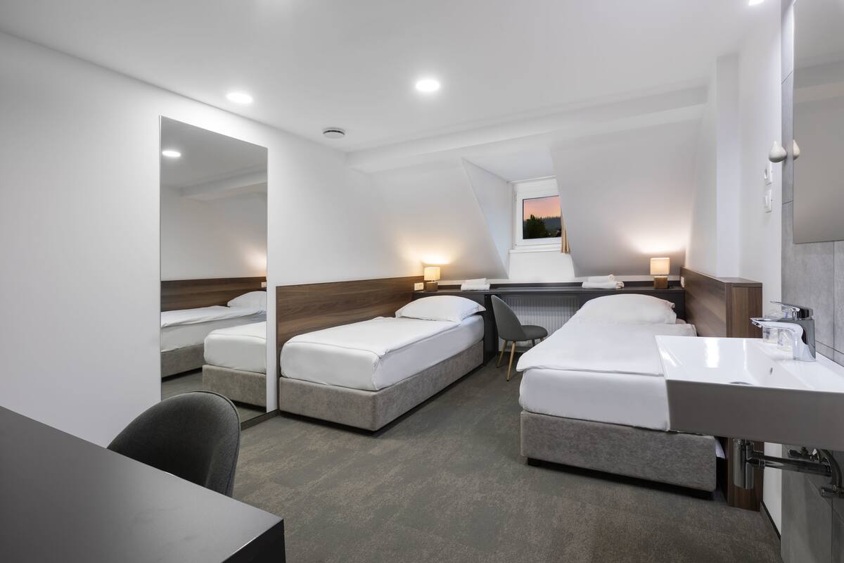 Quarto de hotel moderno com duas camas de solteiro, uma escrivaninha, uma cadeira, um espelho grande e um banheiro com pia.