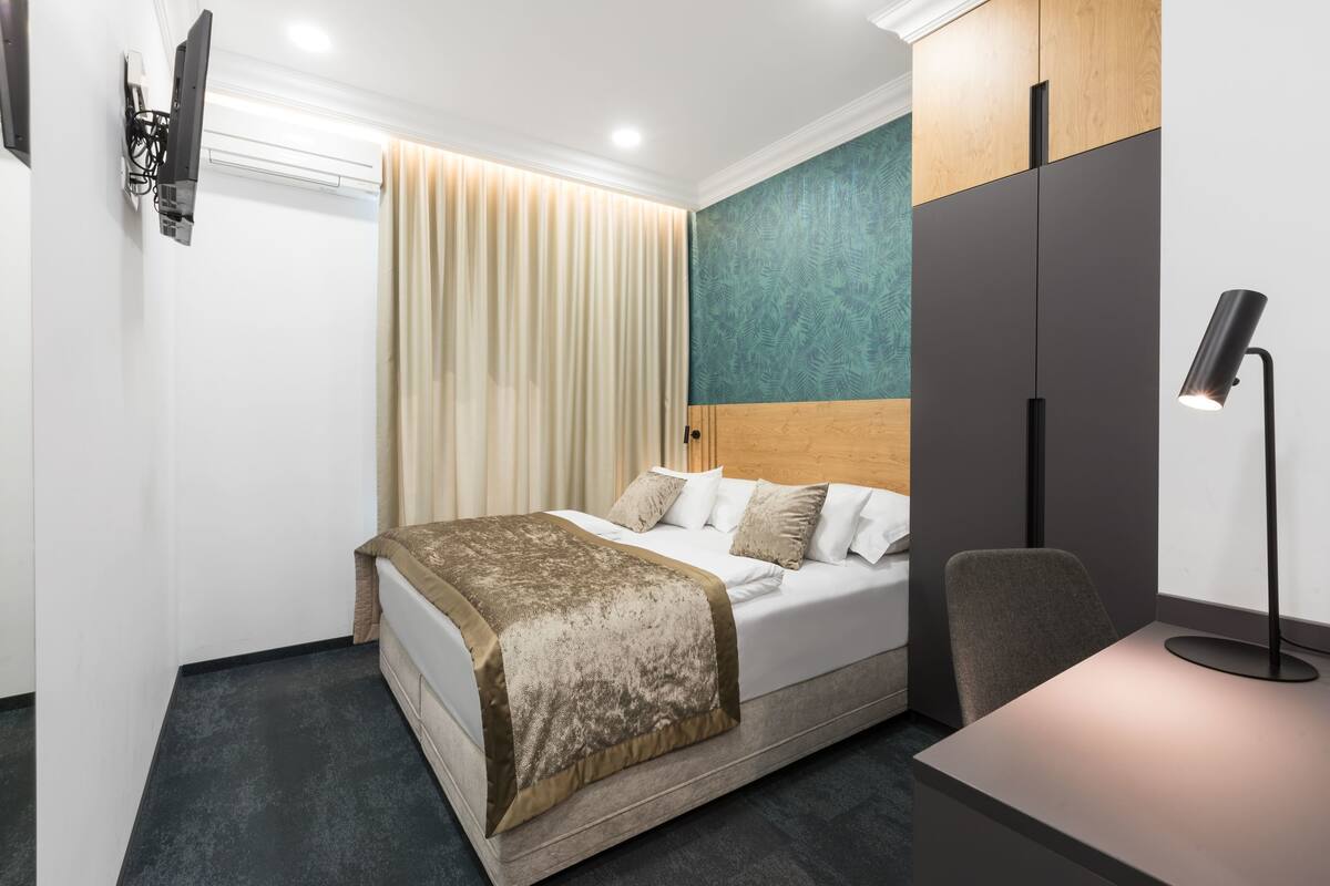 Quarto de hotel moderno com cama, escrivaninha, cadeira, televisão fixada na parede e janela com cortinas.