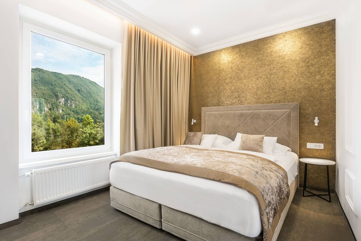 Quarto moderno com uma cama grande, vista para uma floresta pela janela e parede texturizada dourada.