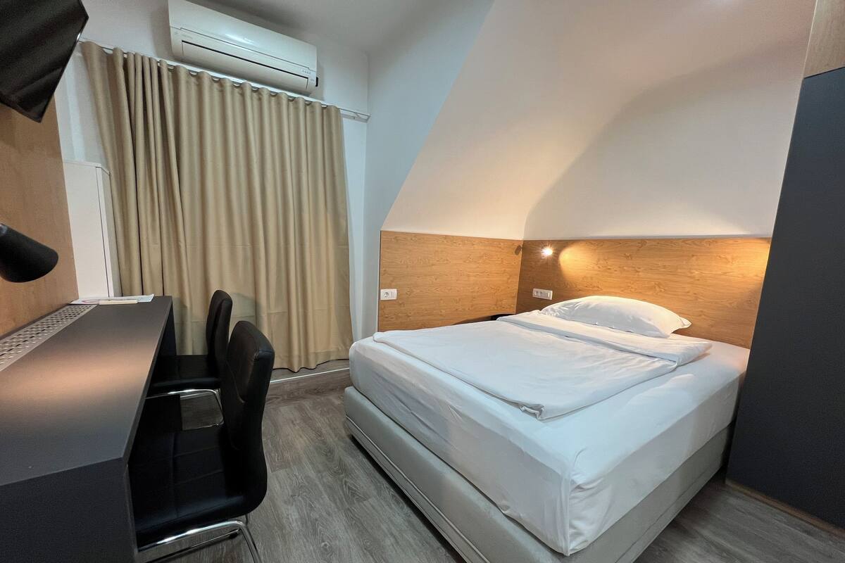 Quarto de hotel com cama, escrivaninha, cadeira, ar condicionado e janela com cortinas.