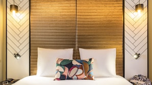 Quarto de hotel com cabeceira de madeira, duas luminárias de apoio, almofada colorida e cama bem arrumada.