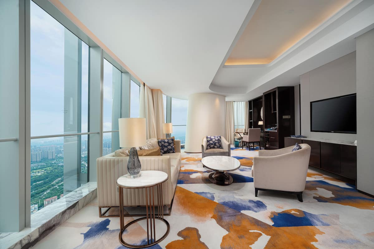Quarto de hotel moderno com uma grande janela, um sofá, uma poltrona, uma televisão e uma mesa de jantar.