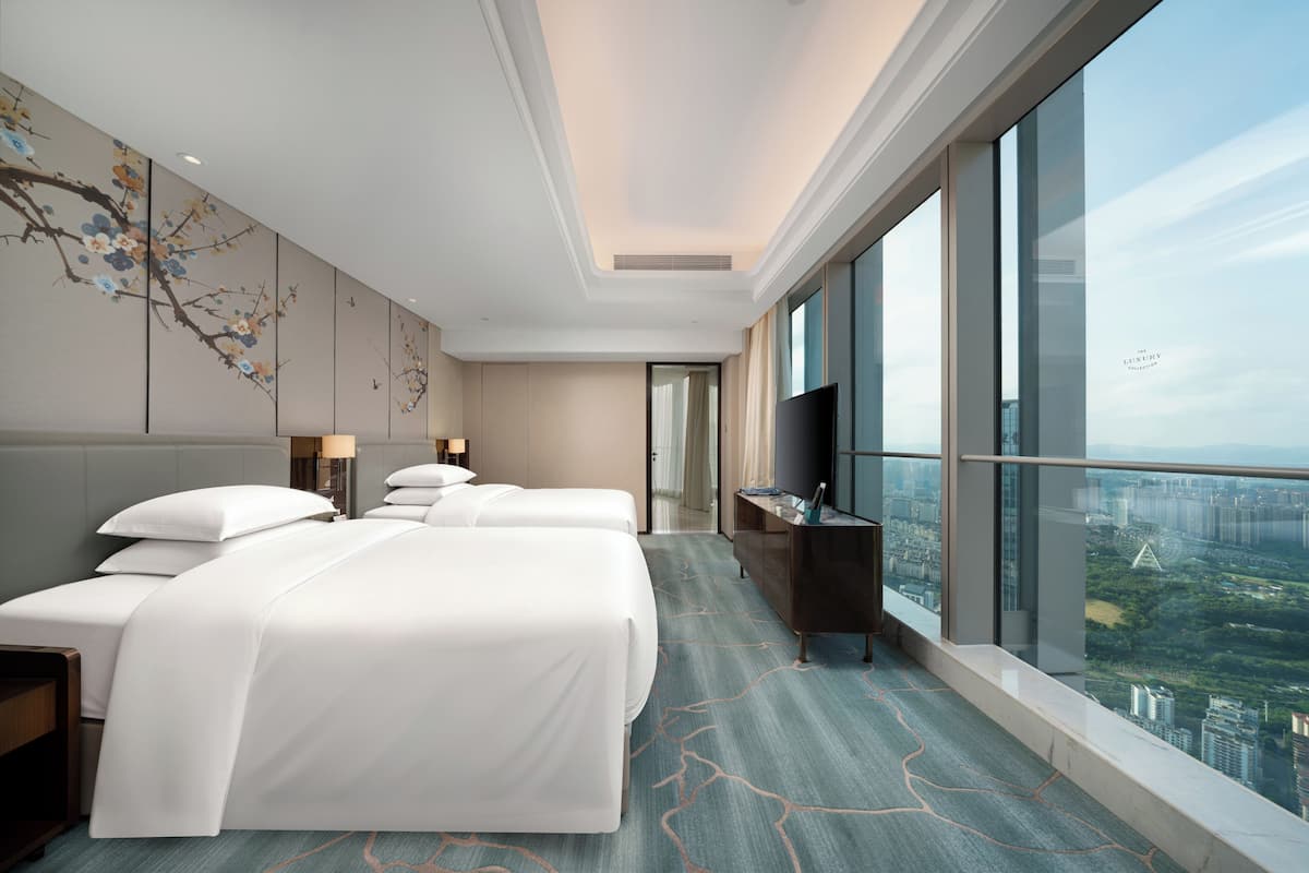 Quarto de hotel com duas camas, uma escrivaninha, uma TV e uma janela grande com vista para a cidade.