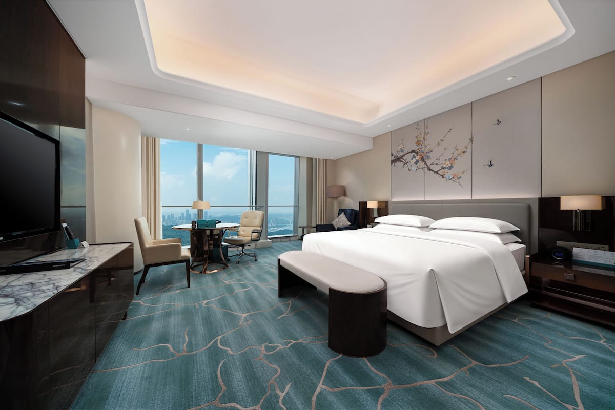 Quarto de hotel com uma cama grande, uma escrivaninha com televisão, uma cadeira, um banco e vista para a cidade.