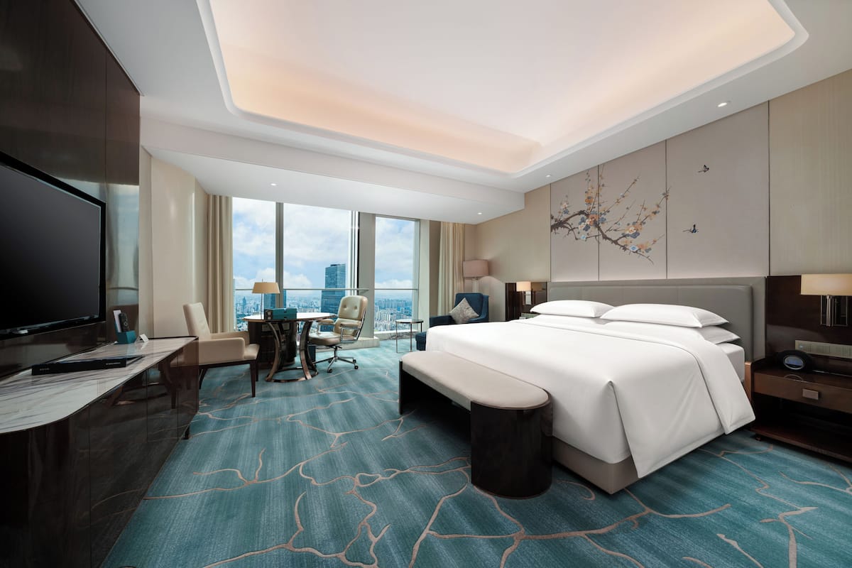Quarto de hotel com uma cama grande, uma escrivaninha com televisão, uma cadeira, uma luminária de mesa e vista para a cidade.