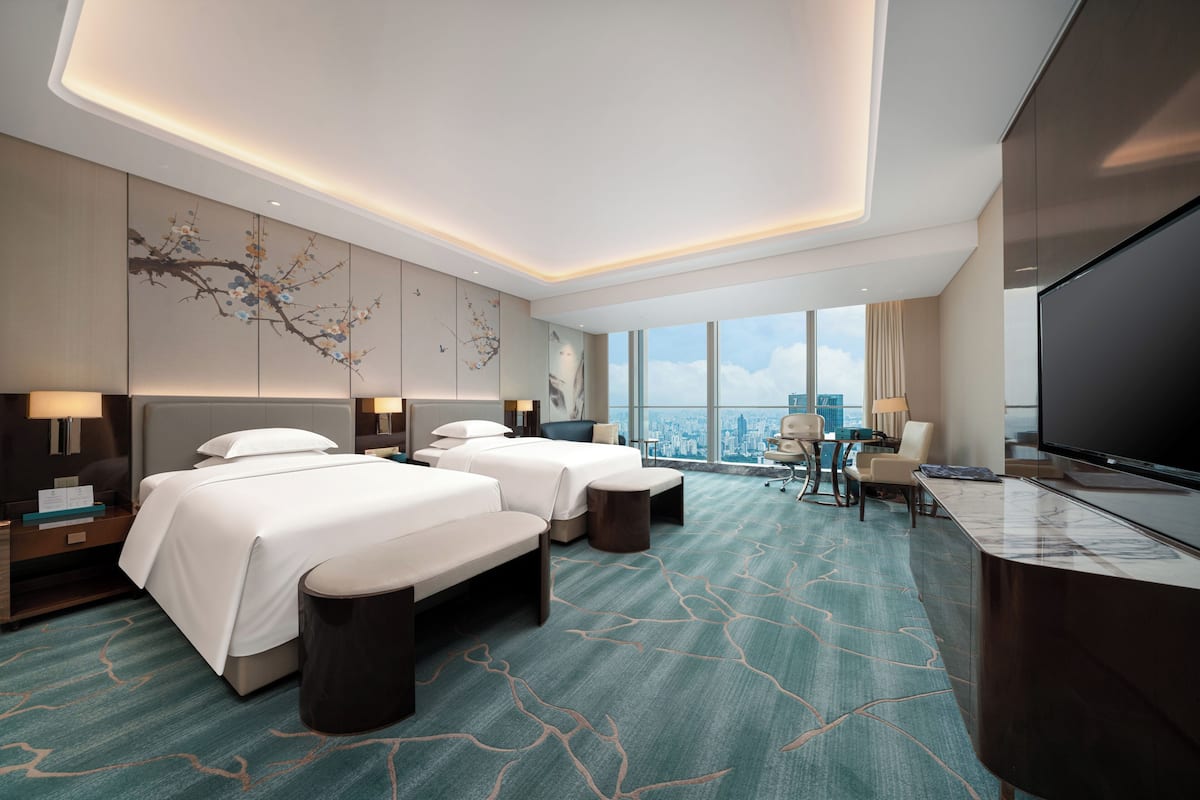 Quarto de hotel com duas camas, uma escrivaninha, televisão e vista para a cidade.