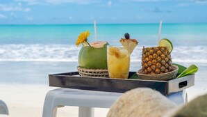 Uma bandeja com três bebidas em uma praia, com céu azul claro e oceano ao fundo.