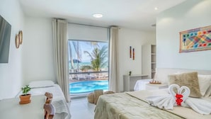 Quarto de hotel com uma cama, uma pequena mesa, uma cadeira, uma planta em vaso e vista para uma piscina e palmeiras.