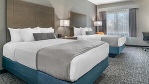 Quarto de hotel com duas camas, uma janela grande com cortinas, televisão de tela plana e uma escrivaninha com luminária.