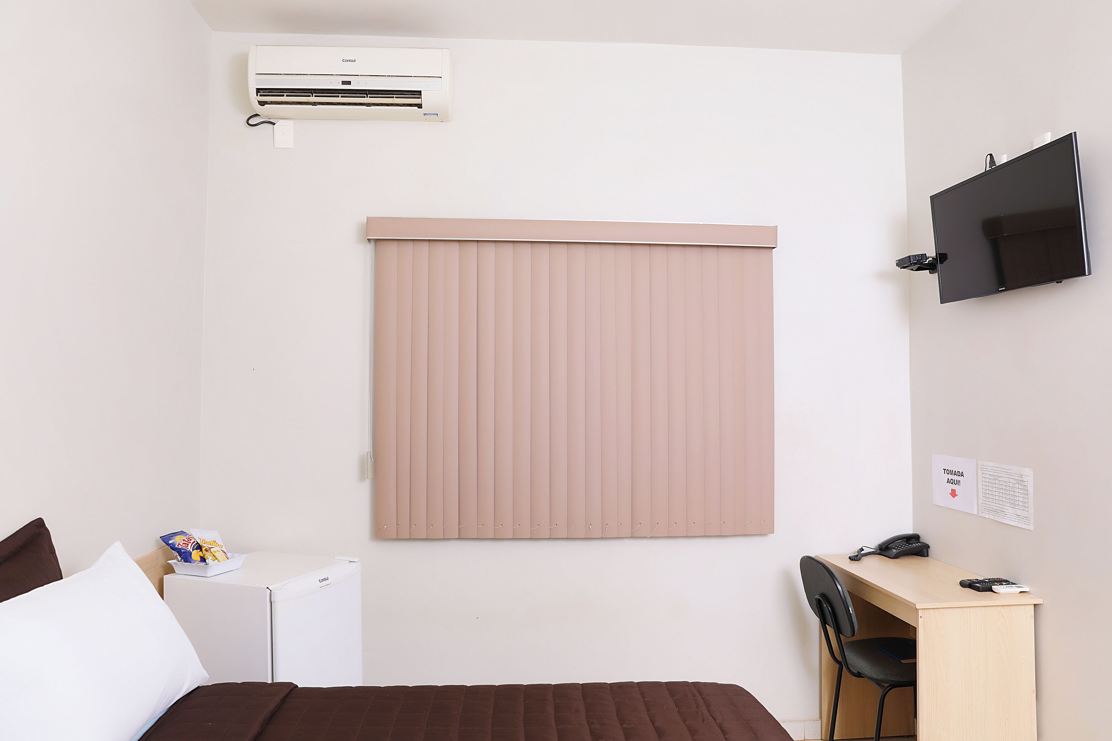 Quarto de hotel com cama, escrivaninha, televisão, ar condicionado e janela com persianas.