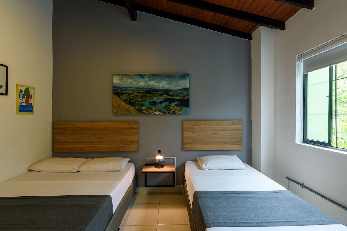 Quarto de hotel com duas camas, uma mesa de cabeceira com abajur, um quadro na parede e uma janela com vista.