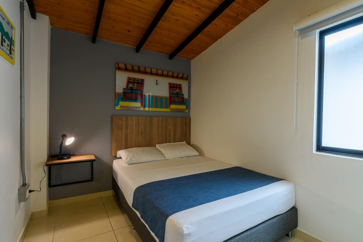 Quarto com cama, mesa de cabeceira, prateleira na parede com obras de arte coloridas e uma janela.