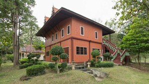 Um edifício tradicional de dois andares com fachada vermelha, uma escadaria que leva ao andar superior e um jardim bem cuidado com arbustos e árvores podados.